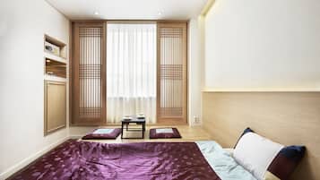 Quarto com cama, almofadas, uma mesinha e janela com persianas.