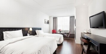 Quarto de hotel com duas camas, uma escrivaninha, uma cadeira, uma televisão e uma janela com cortinas.
