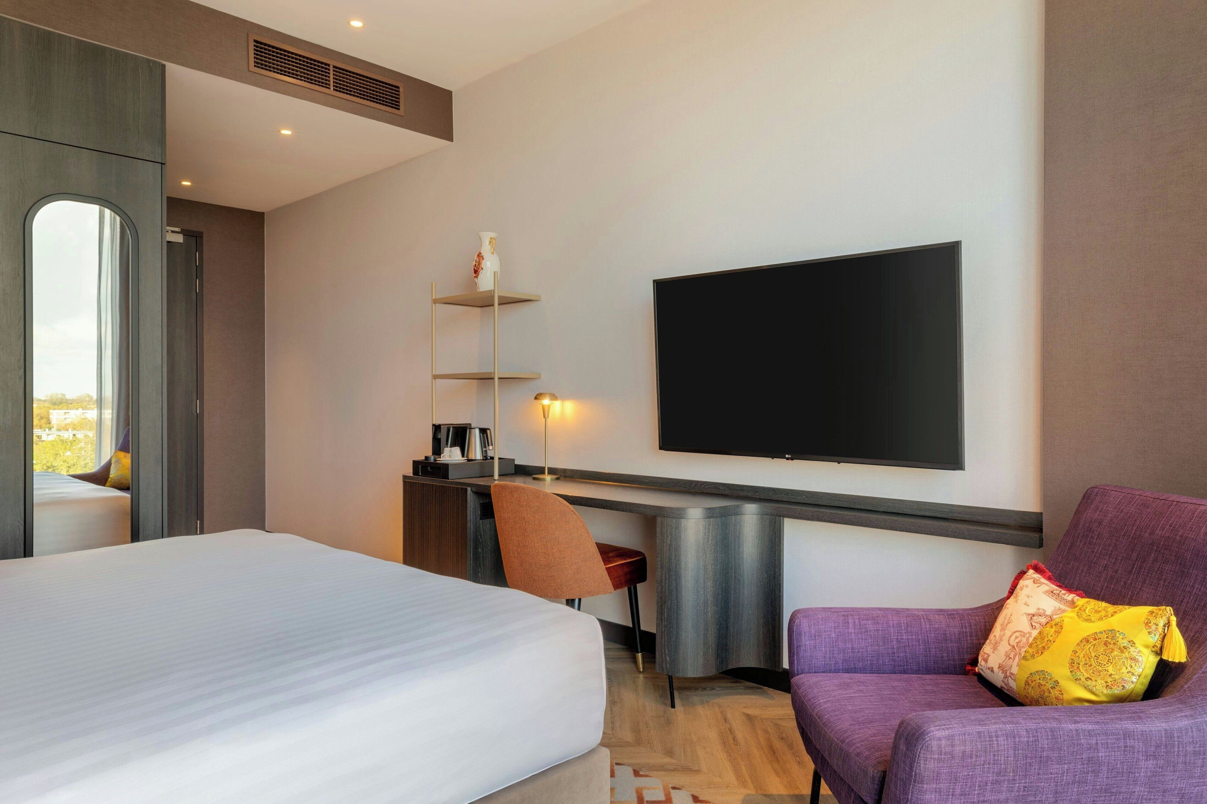 Quarto de hotel com cama, escrivaninha, cadeira, televisão e janela.