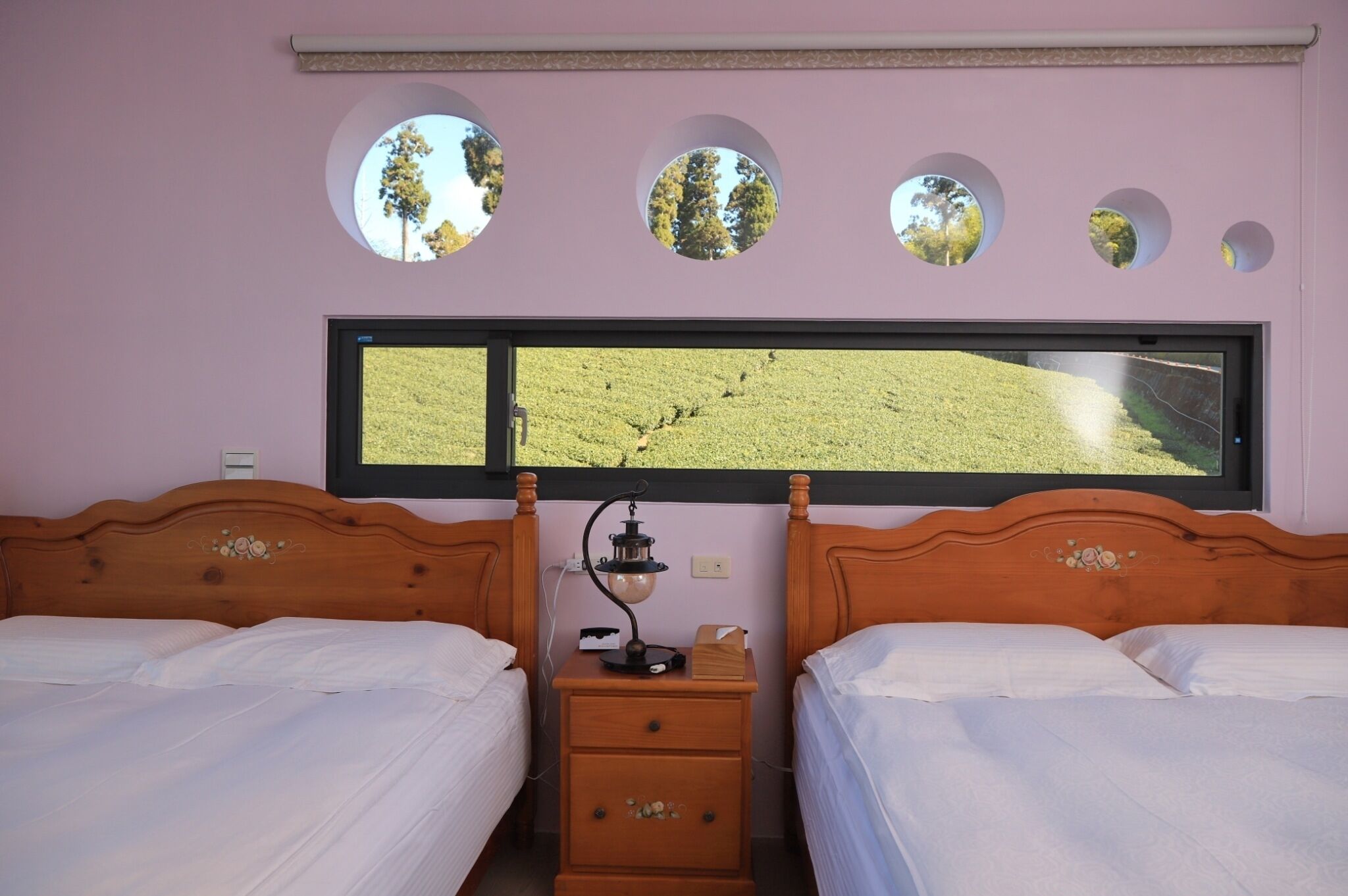 Quarto com duas camas, mesa de cabeceira com abajur e janela com vista para um campo gramado.