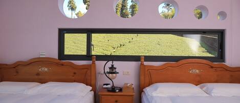 Quarto com duas camas, mesa de cabeceira com abajur e janela com vista para um campo gramado.