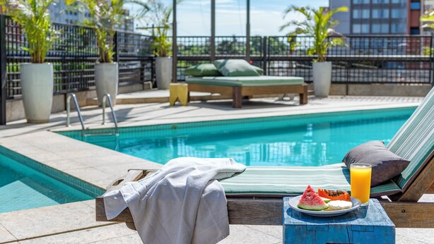 Uma área de piscina no terraço com uma espreguiçadeira, uma mesa pequena com uma travessa de fruta e um copo de suco de laranja, além de uma cama com almofadas.