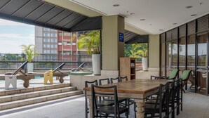Uma área de refeições ao ar livre com uma grande mesa de madeira e cadeiras, rodeada por um alpendre coberto com vista para um edifício e palmeiras.