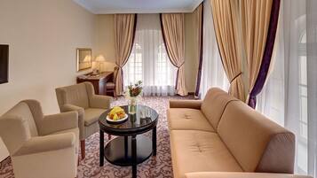 Quarto de hotel com um sofá bege, duas poltronas, uma mesa redonda pequena com uma fruteira, um abajur e um quadro na parede.