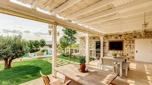 Um pátio externo com mesa e cadeiras de madeira, um muro de pedra e vista para uma piscina e jardim.