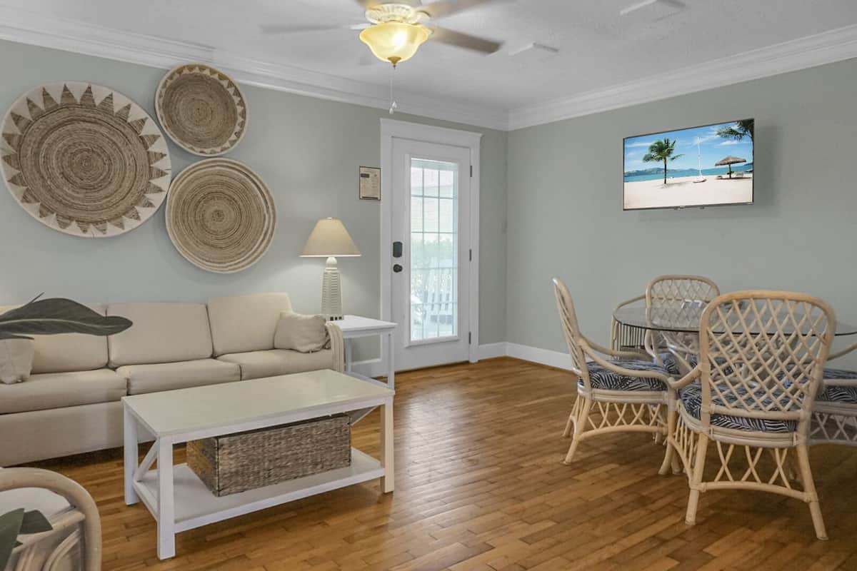 Sala de estar com sofá, mesa de centro e cadeiras de vime. Há um ventilador de teto, um abajur e um quadro com uma paisagem de praia na parede.
