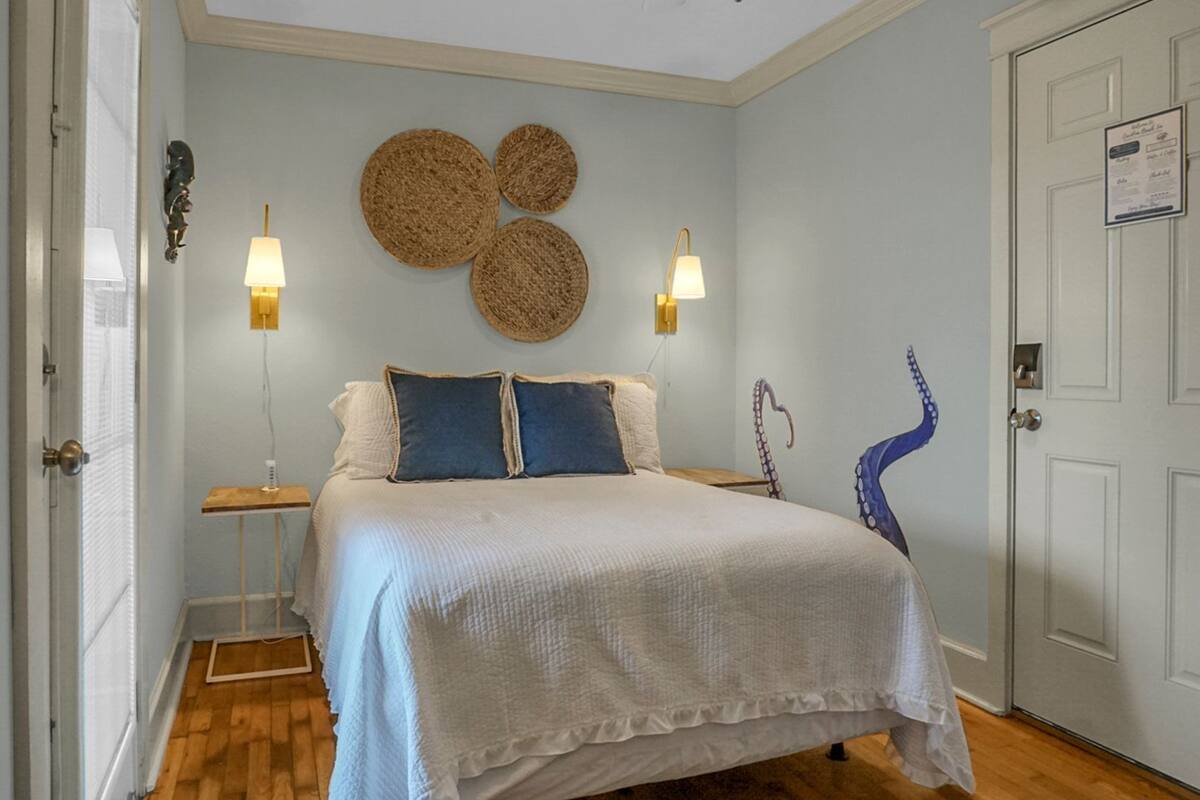 Quarto com uma cama, duas luminárias de cabeceira, um objeto decorativo na parede e uma porta com uma placa.