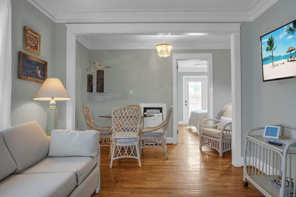 Uma sala de estar ampla com piso de madeira, um sofá branco, cadeiras de vime, uma mesa de jantar e um berço para bebê.