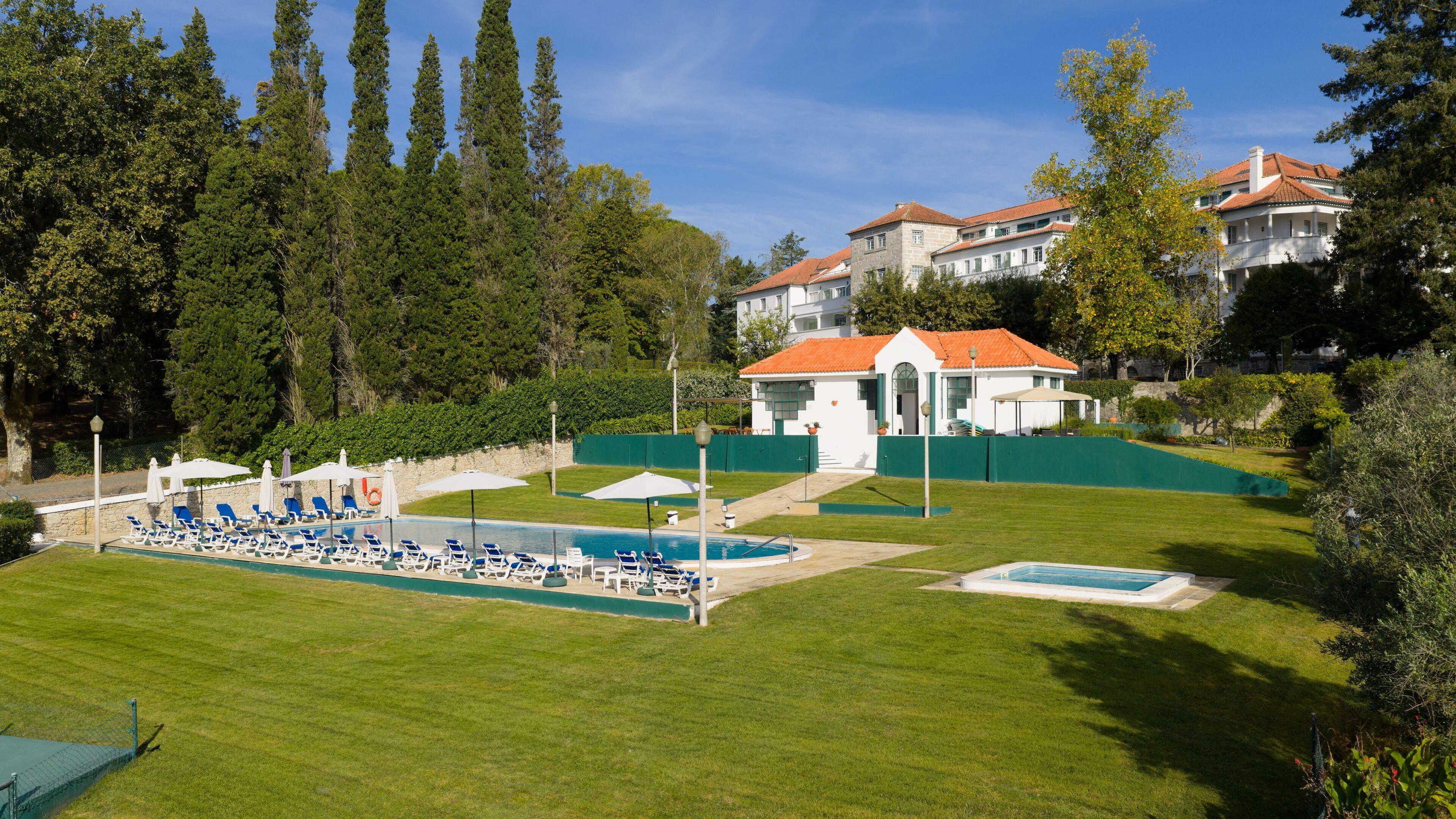 Uma área de piscina com espreguiçadeiras e guarda-sóis, rodeada por gramados bem cuidados e árvores.