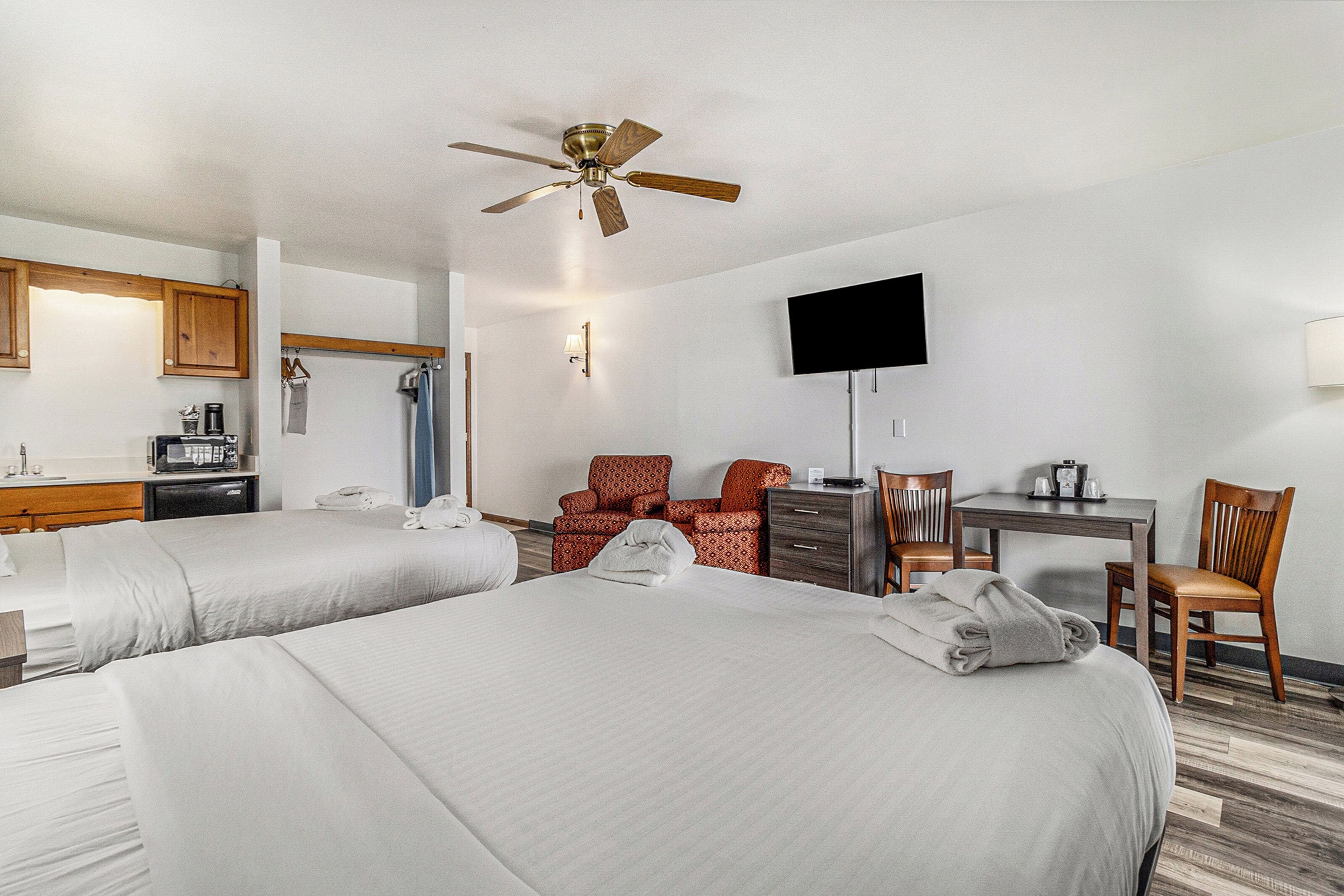Quarto de hotel com duas camas, uma escrivaninha, uma cadeira, uma televisão e um ventilador de teto.