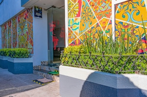 Fachada de edifício com padrões geométricos coloridos, cerca de metal e um pequeno jardim com plantas verdes.