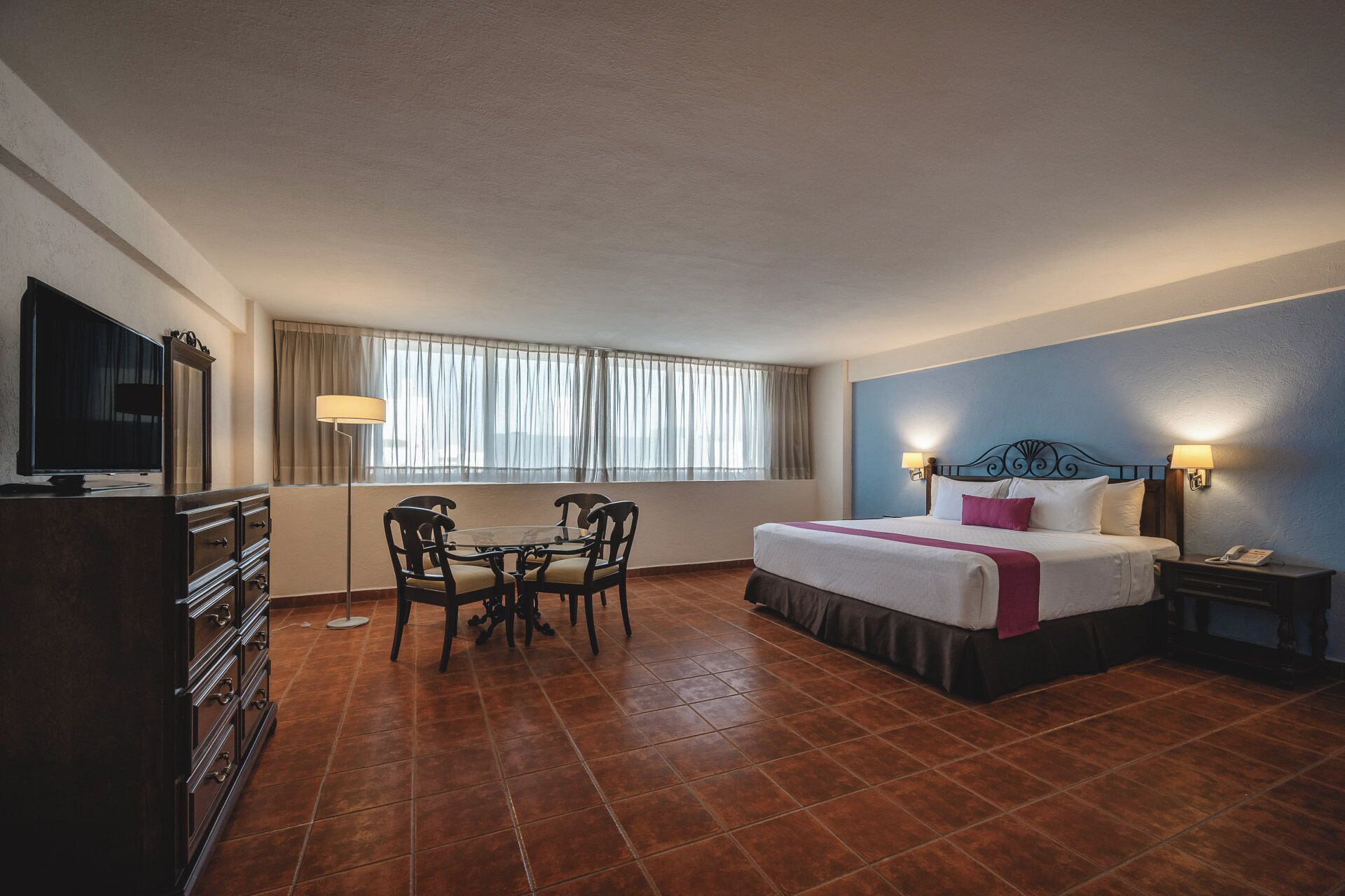 Quarto de hotel com uma cama, uma escrivaninha com candeeiro, uma televisão e uma mesa de jantar com cadeiras.