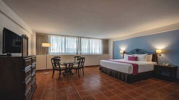 Quarto de hotel com uma cama, uma escrivaninha com candeeiro, uma televisão e uma mesa de jantar com cadeiras.