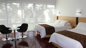 Quarto de hotel com duas camas, uma mesa de vidro com cadeiras e amplas janelas com cortinas finas.