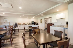 Uma sala de jantar espaçosa, com mesas e cadeiras de madeira, uma cozinha com armários brancos e um teto com iluminação embutida.