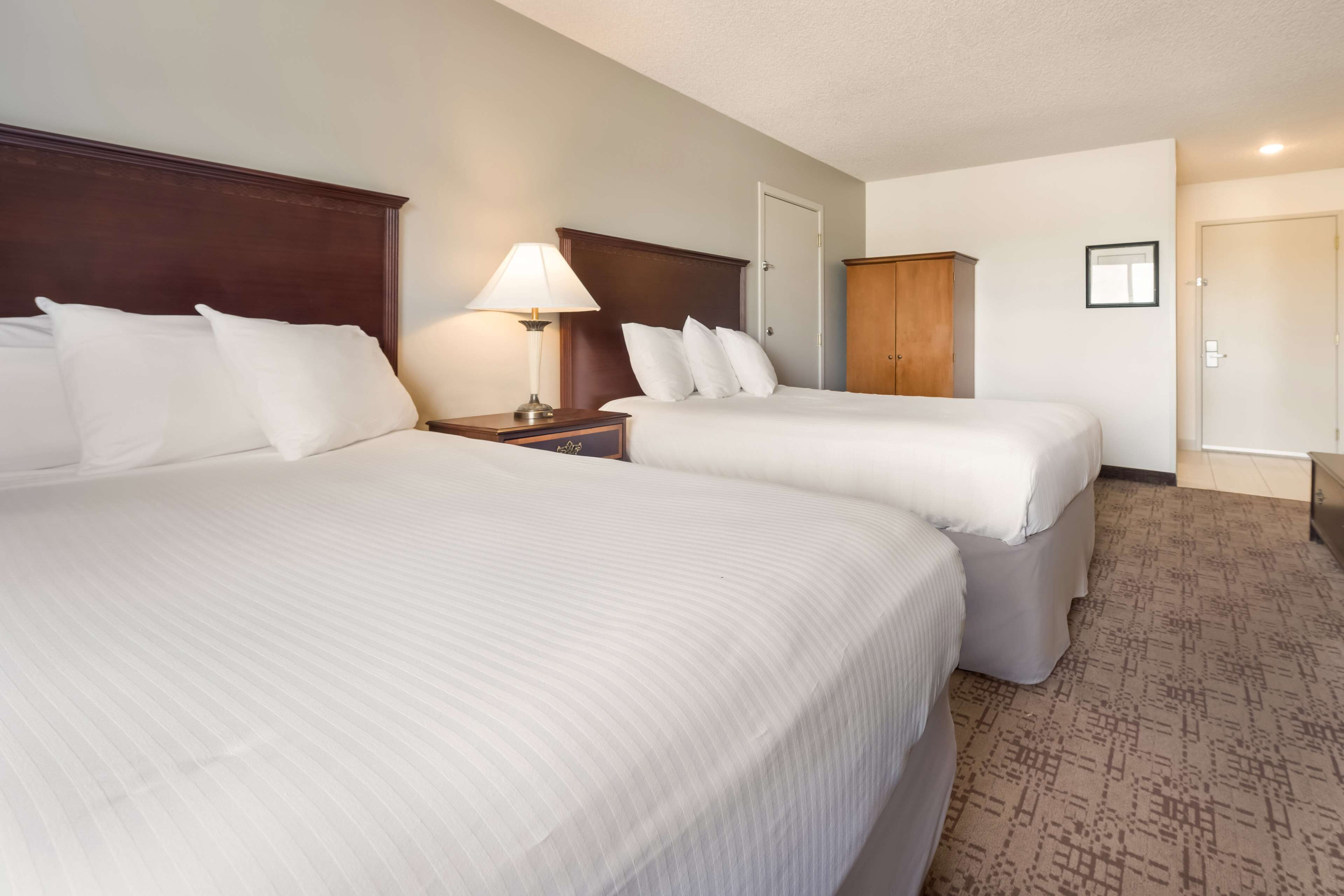 Quarto de hotel com duas camas, uma mesa de cabeceira com abajur, um guarda-roupa de madeira e piso coberto por carpete.