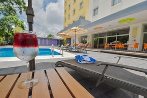 Uma área na beira da piscina, com um copo de bebida vermelha, cadeiras de descanso, guarda-sóis e um prédio ao fundo.