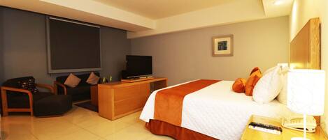 Quarto de hotel com cama, televisão, escrivaninha, cadeira e sofá.