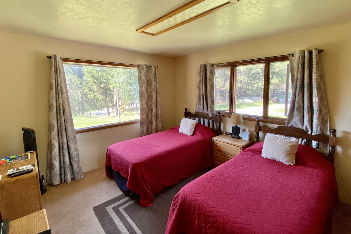 Quarto de hotel com duas camas, mesa de cabeceira com abajur, relógio e janela com cortinas.