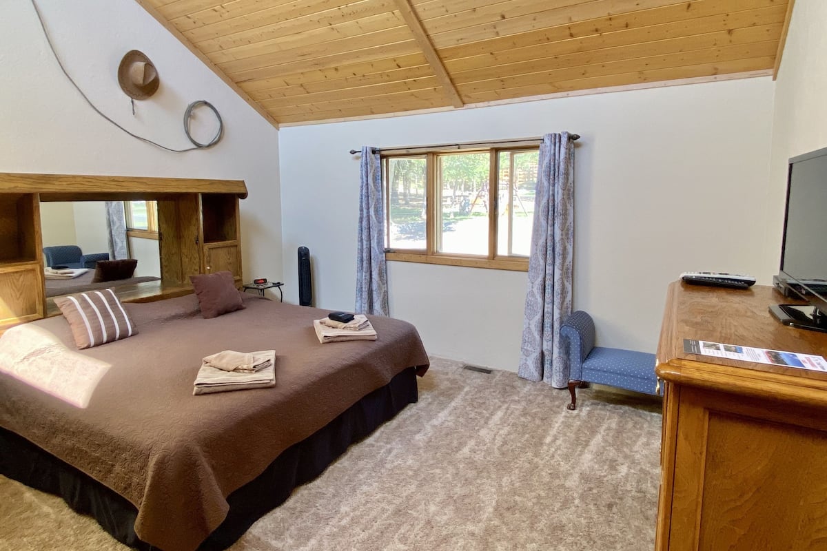 Quarto com teto de madeira, cama com colcha marrom, escrivaninha com televisão e janela com cortinas.