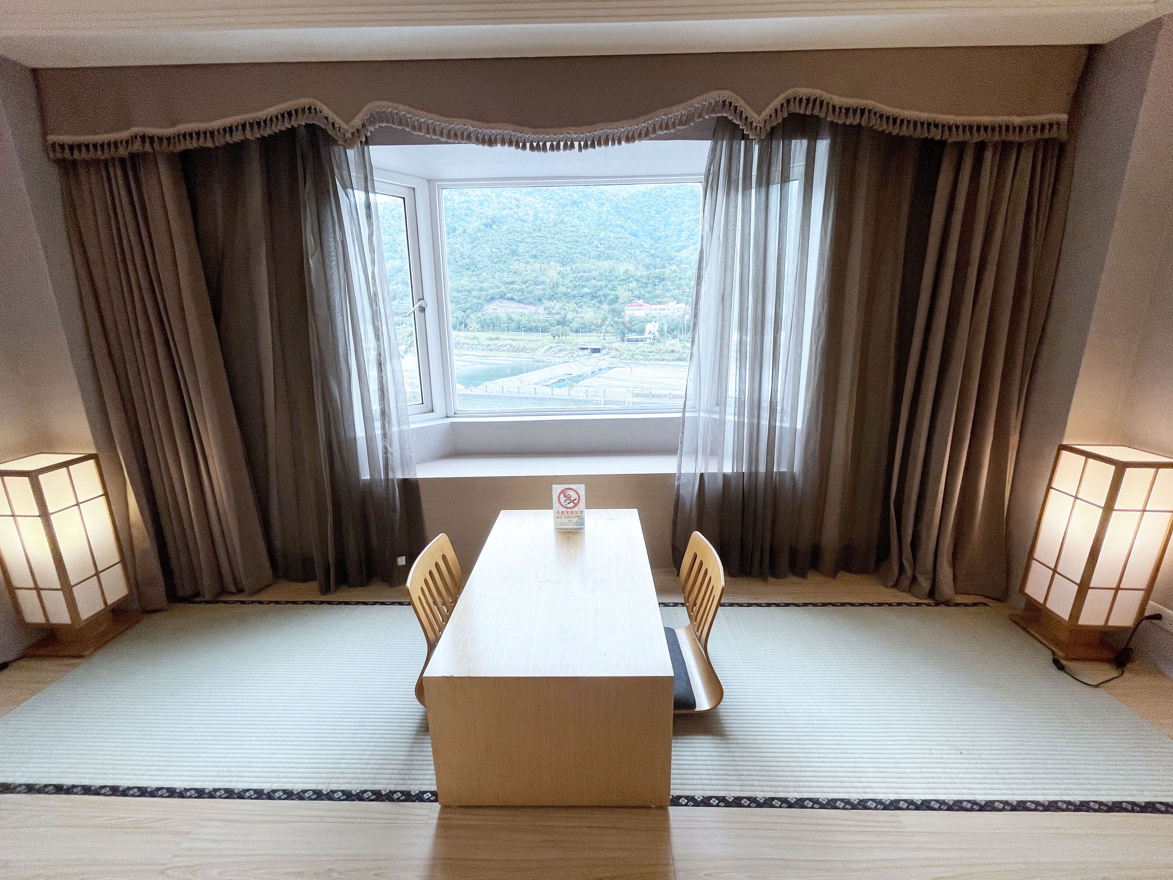 Quarto de hotel com mesa de jantar, cadeiras, vista pela janela e duas luminárias.