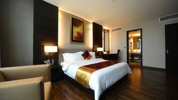 Quarto de hotel com uma cama grande, área de estar com uma poltrona, escrivaninha e espelho.