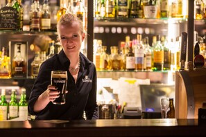 Um bartender segurando um copo de cerveja Guinness em um bar, com diversas garrafas de bebidas alcoólicas ao fundo.