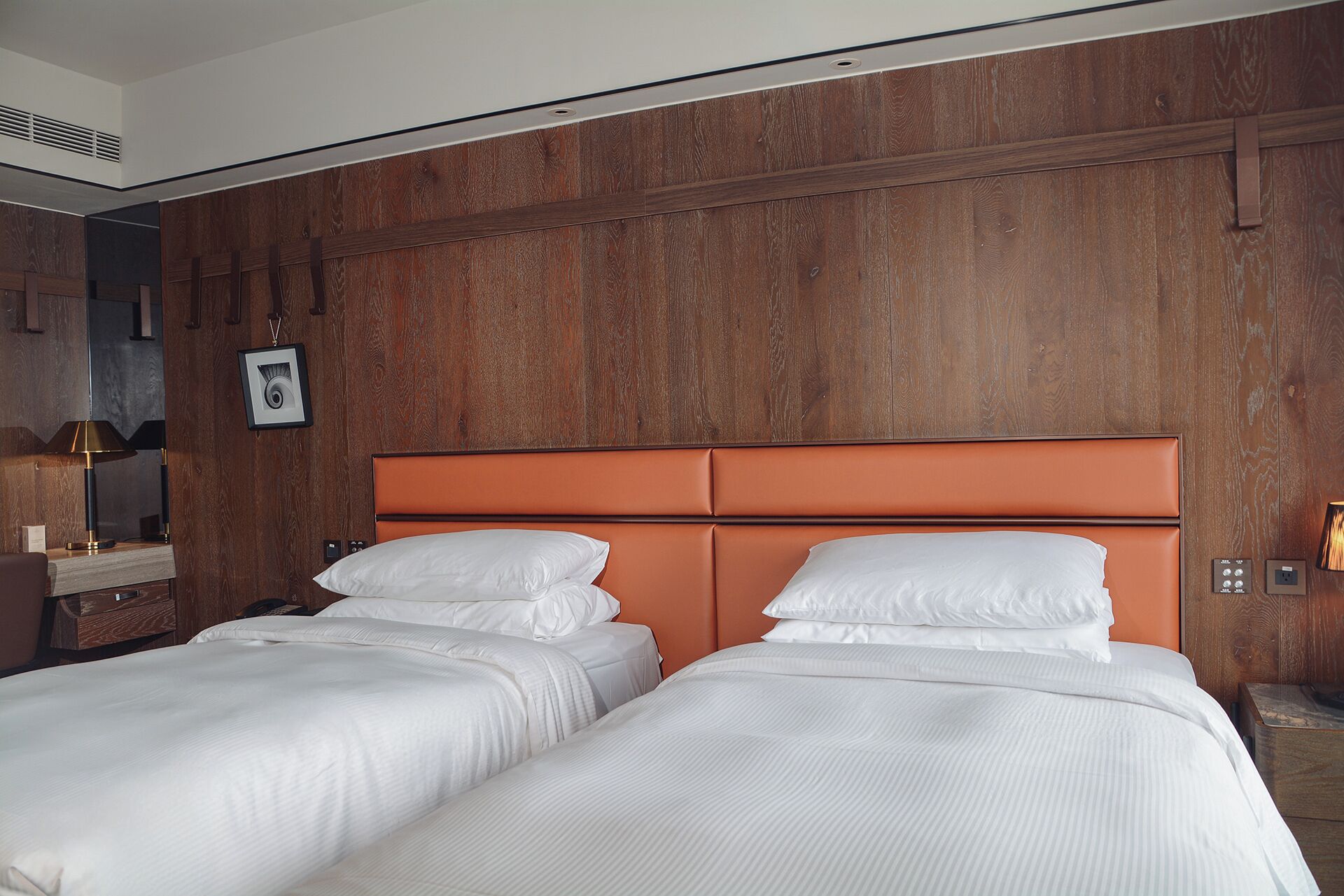 Quarto de hotel com duas camas, paredes de painel de madeira, um quadro emoldurado e uma mesa de cabeceira com abajur.