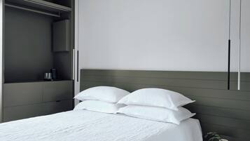 Quarto moderno com cama, roupa de cama branca, mesa de cabeceira com um livro e uma planta, e guarda-roupa com espelho.