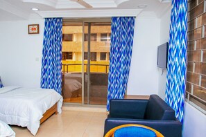 Quarto de hotel com cama, sofá, televisão e varanda com cortinas estampadas.