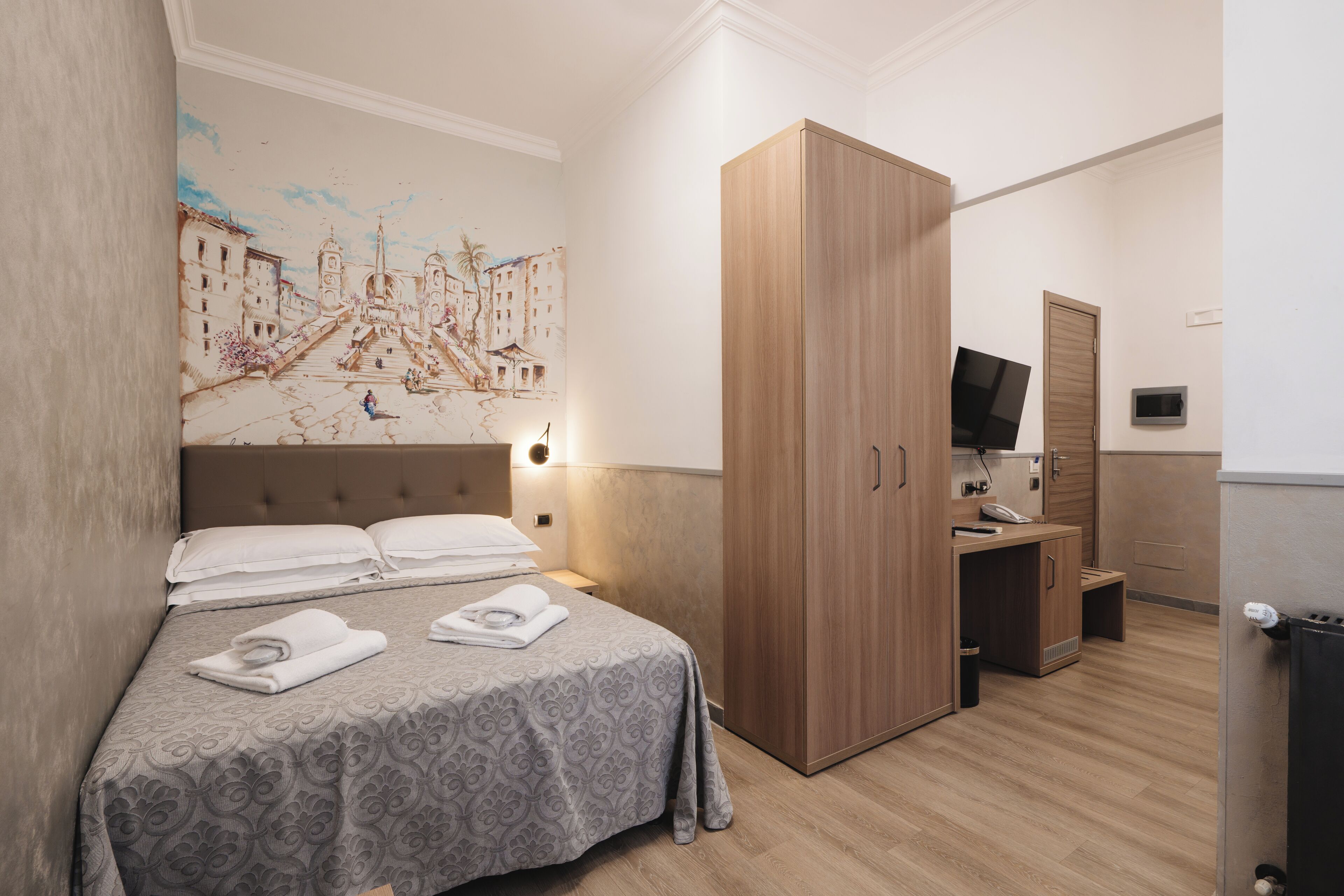 Quarto de hotel com uma cama, um guarda-roupa, uma secretária com um computador e um quadro na parede.
