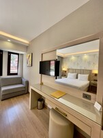 Quarto de hotel com cama, sofá, escrivaninha com espelho, televisão e janela com cortinas.