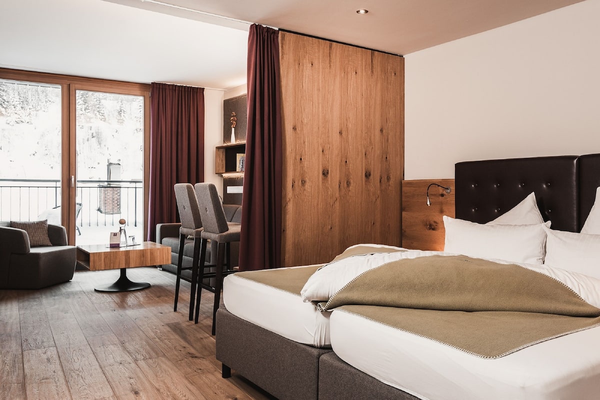 Quarto de hotel moderno com uma cama grande, área de estar com sofá e cadeiras, parede de destaque em madeira e vista através de amplas janelas.