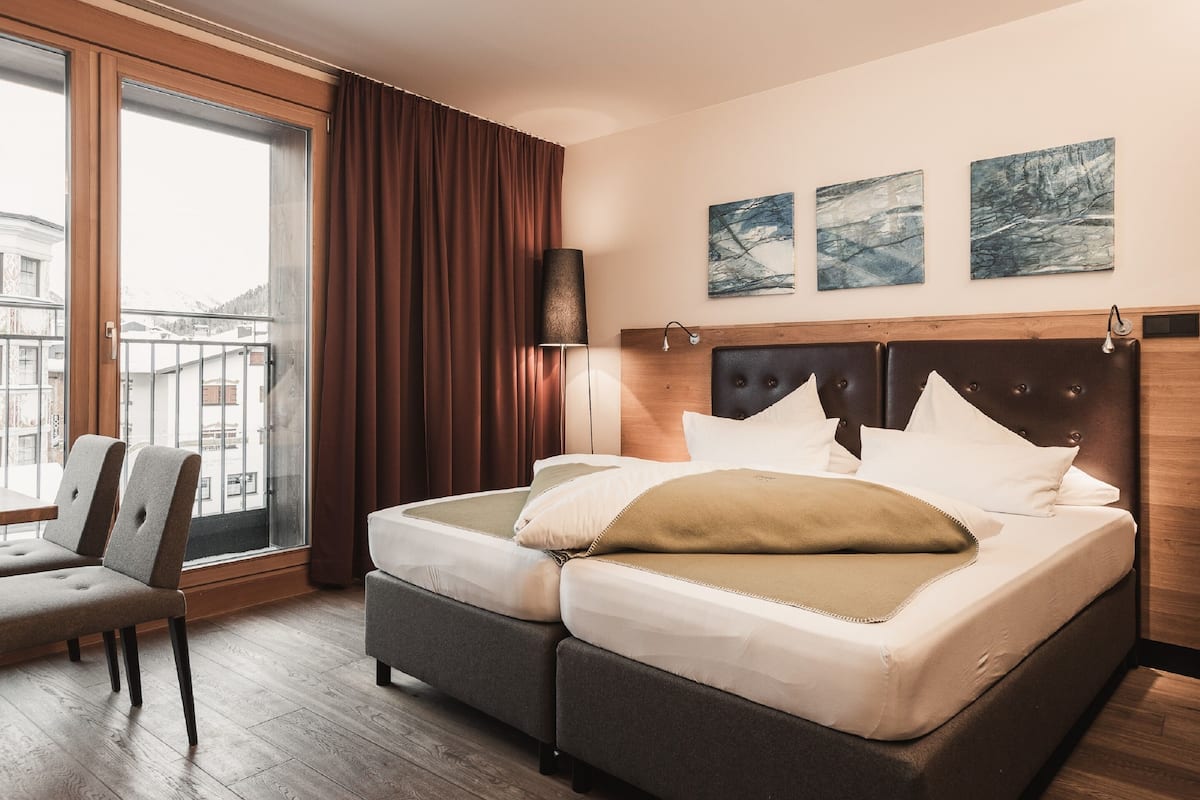 Quarto de hotel com uma cama grande, duas cadeiras, uma janela com cortinas e obras de arte na parede.