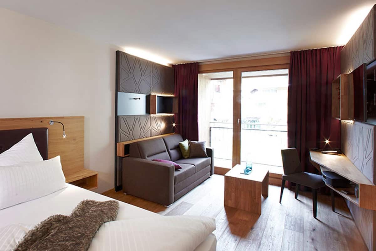 Quarto de hotel moderno com uma cama grande, um sofá, uma escrivaninha e uma varanda com cortinas vermelhas.