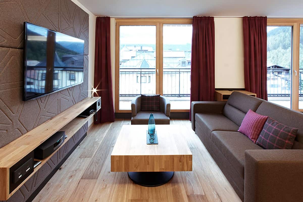 Uma sala de estar moderna com uma televisão de tela plana fixada em uma parede texturizada, uma mesa de centro de madeira, um sofá e uma poltrona. Janelas amplas com cortinas oferecem vista para edifícios e colinas.