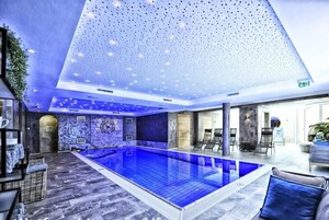 Piscina interior moderna com chão e teto azulejados em azul, rodeada por espreguiçadeiras e um espaço de estar.