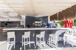 Uma área de bar ao ar livre com balcão, cadeiras brancas e uma cozinha ao fundo.
