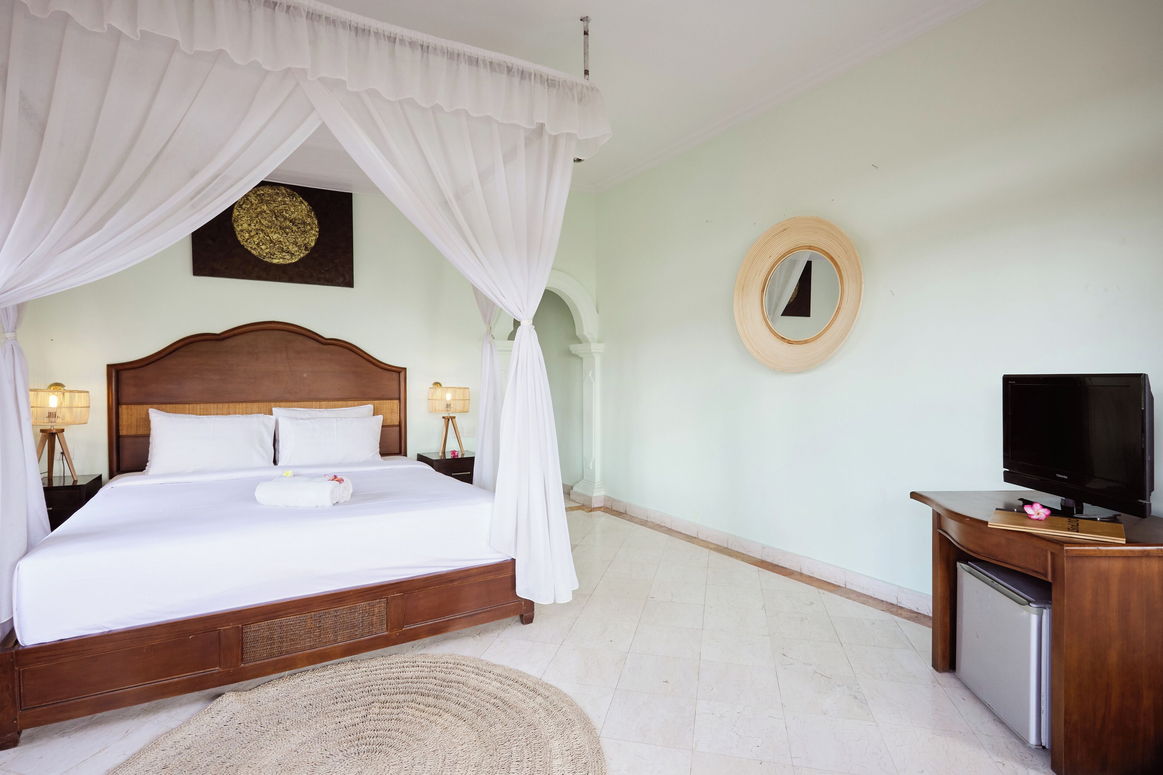 Quarto com cama, televisão, mesa de cabeceira, abajur, espelho e um objeto decorativo na parede.