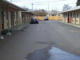 Um motel com placa de escritório em formato de 'C', estacionamento e cerca de madeira ao fundo.