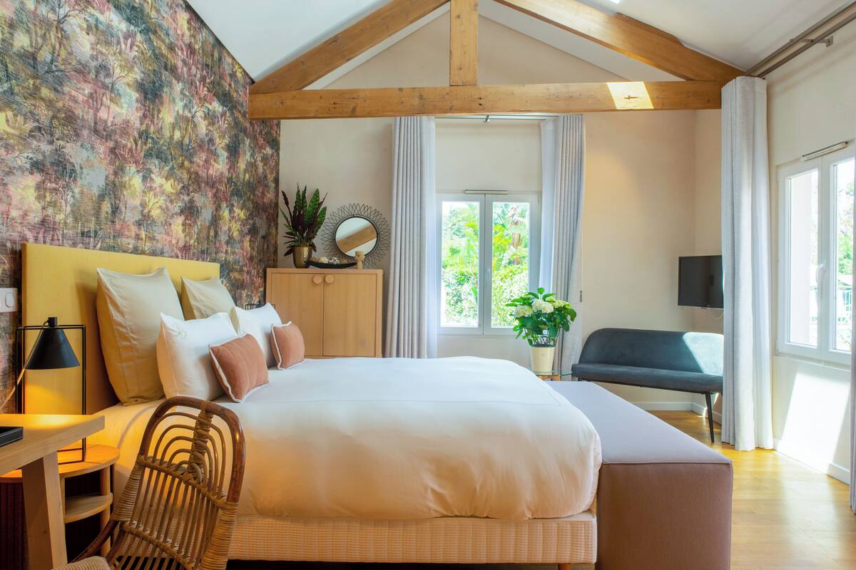 Quarto com uma cama grande, cabeceira de madeira, mesa de cabeceira, uma cadeira, televisão e janela com cortinas.