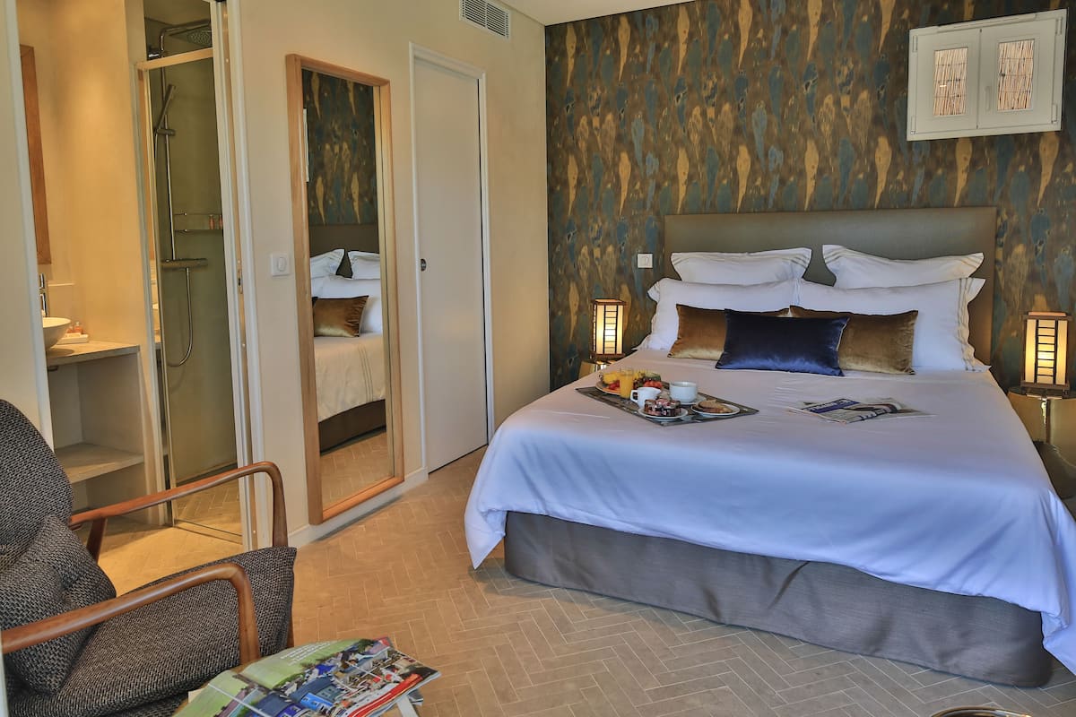 Quarto de hotel com cama, poltrona, mesa de cabeceira, espelho e área de chuveiro.