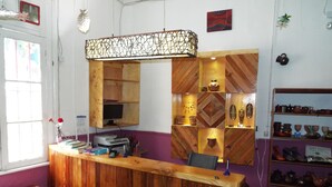 Um balcão de atendimento com acabamento em madeira, um monitor de computador e uma impressora. Há prateleiras com objetos decorativos e uma janela com vista para o exterior.
