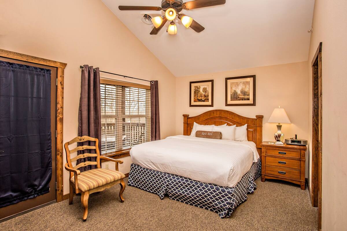 Quarto com cama grande, cadeira de madeira, mesa de cabeceira, luminária e ventilador de teto.
