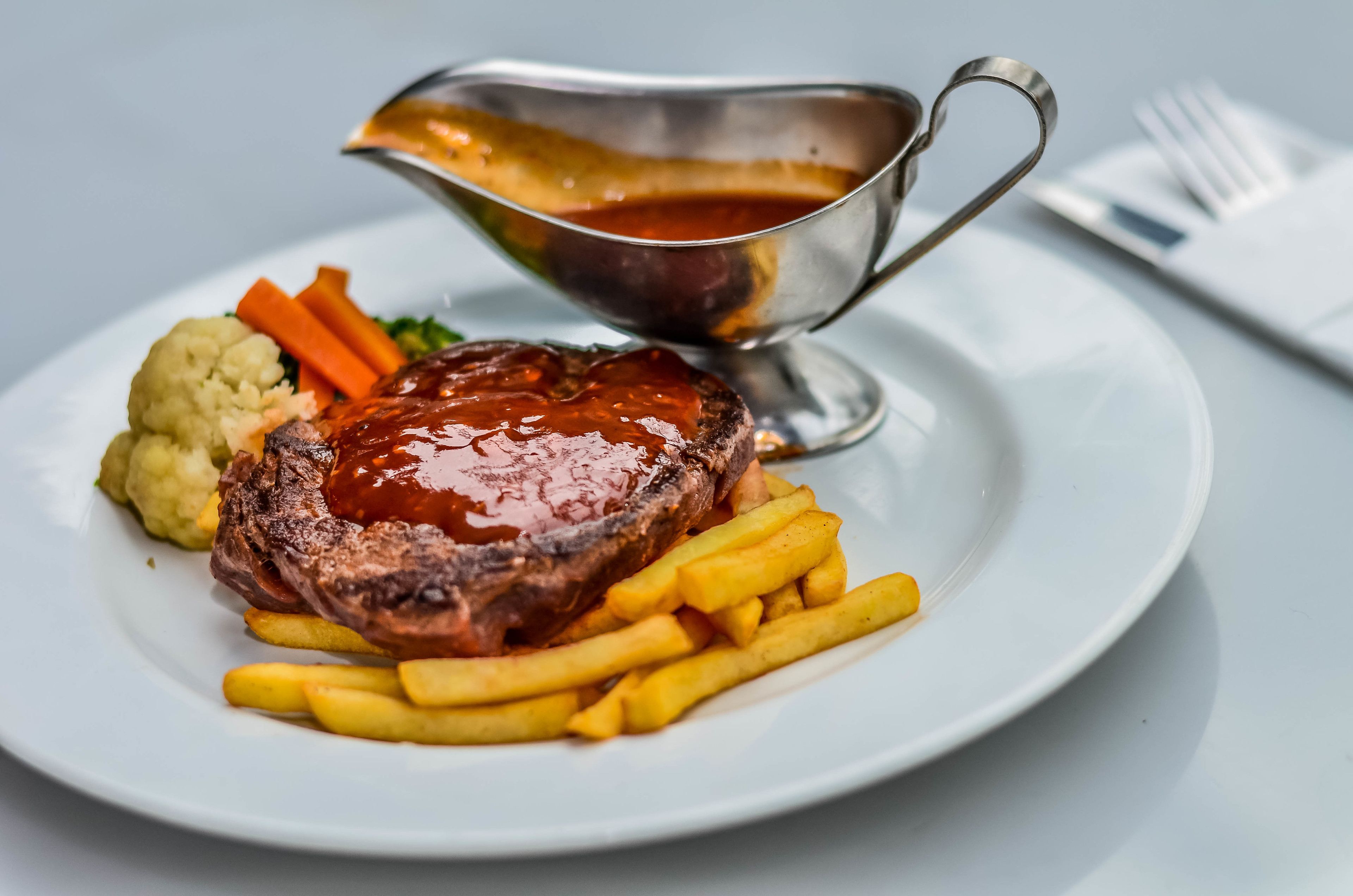 Um prato com bife, batatas fritas e legumes, e um regador de molho.
