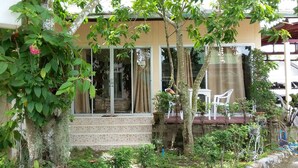 Uma casa com escadaria de pedra que leva a uma porta de vidro, rodeada por vegetação e uma pequena área externa com cadeiras.