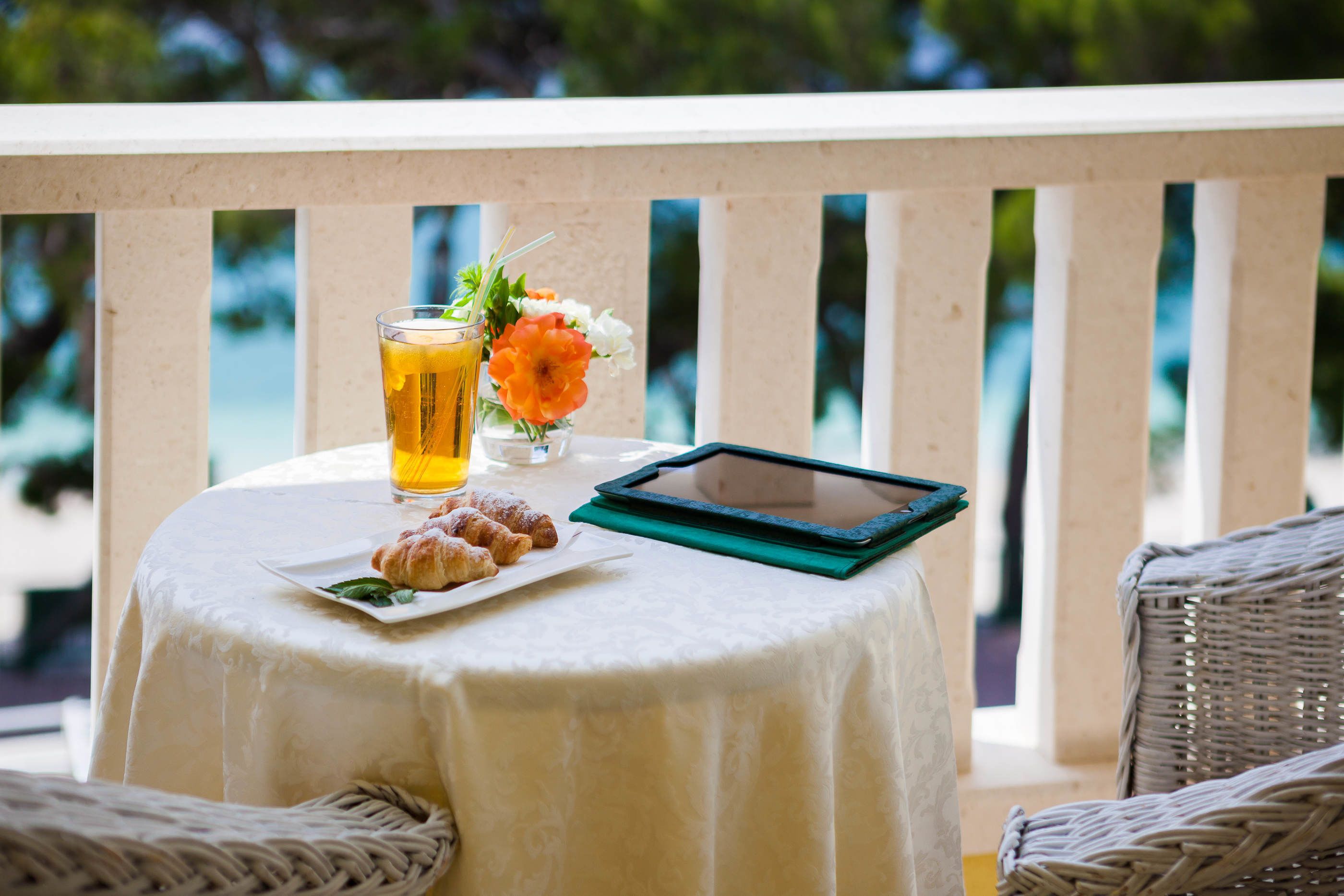 Uma mesa redonda com toalha branca, um copo de chá gelado, um prato com croissants, um pequeno vaso com flores e um tablet sobre a mesa.