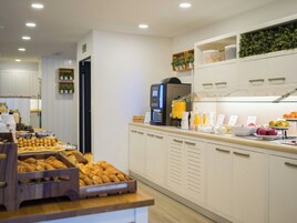 Uma área de buffet com diversos alimentos, uma máquina de café e uma vitrine de vidro com frutas.
