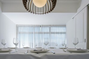 Uma sala de jantar com mesa posta para seis pessoas, um lustre moderno e uma janela grande com cortinas leves.