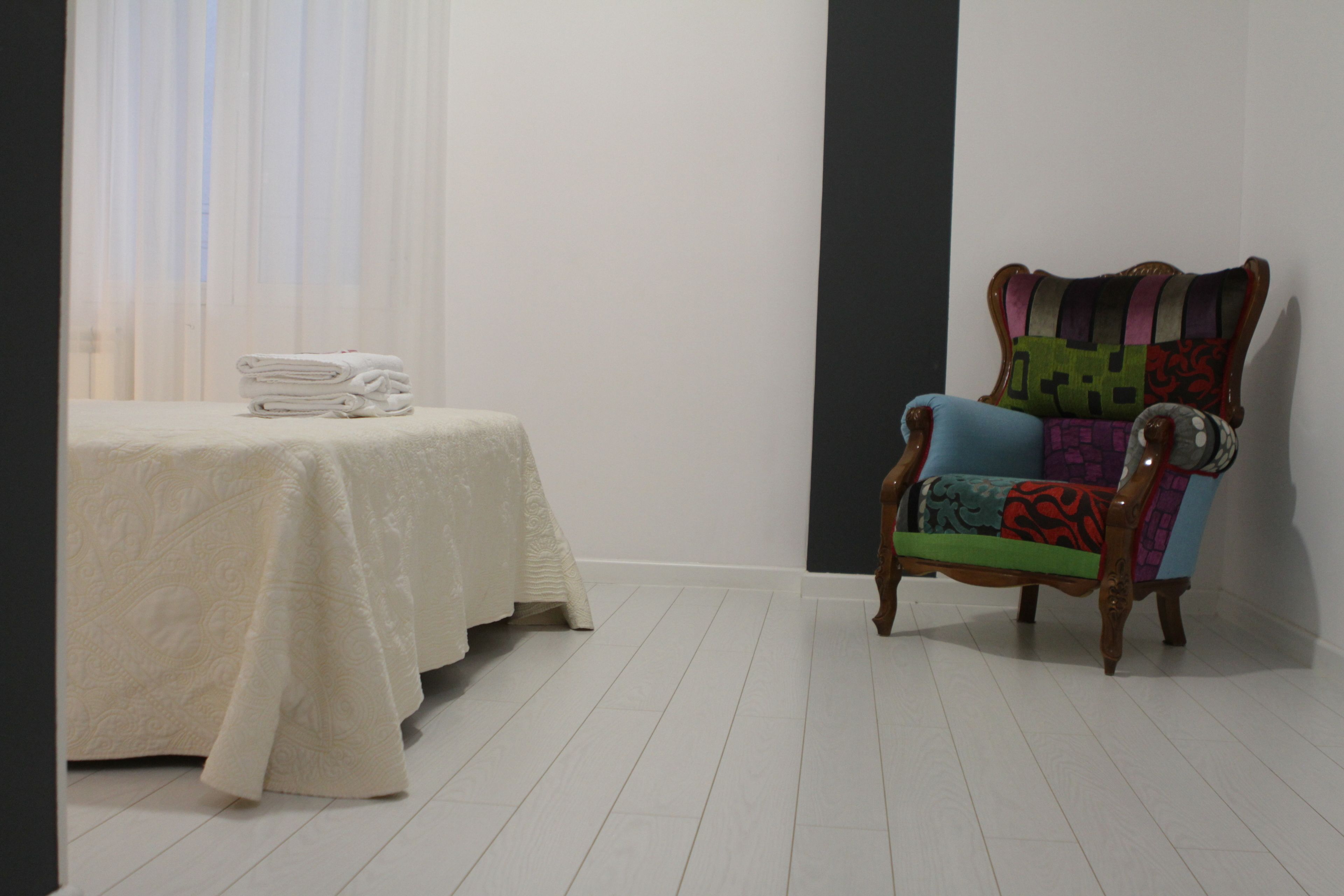 Quarto com cama coberta por um lençol branco, uma cadeira com almofadas coloridas e uma mesinha com toalhas dobradas.